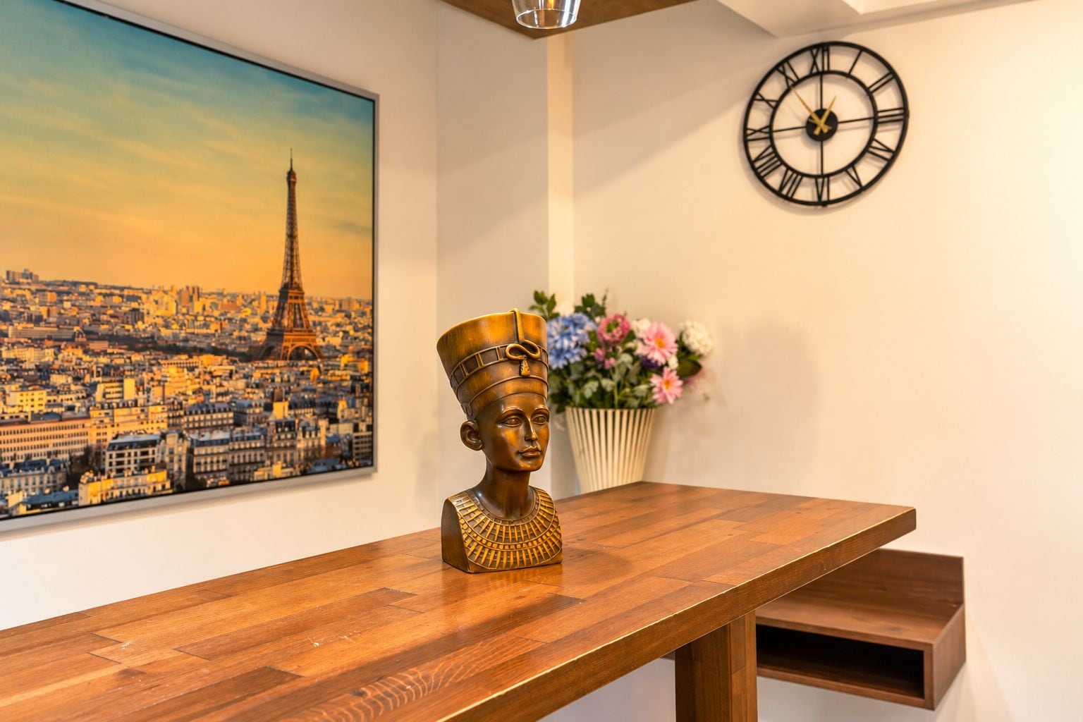 Uma mesa de madeira com um busto decorativo, um relógio de parede, uma pintura emoldurada de uma vista urbana e um vaso com flores.
