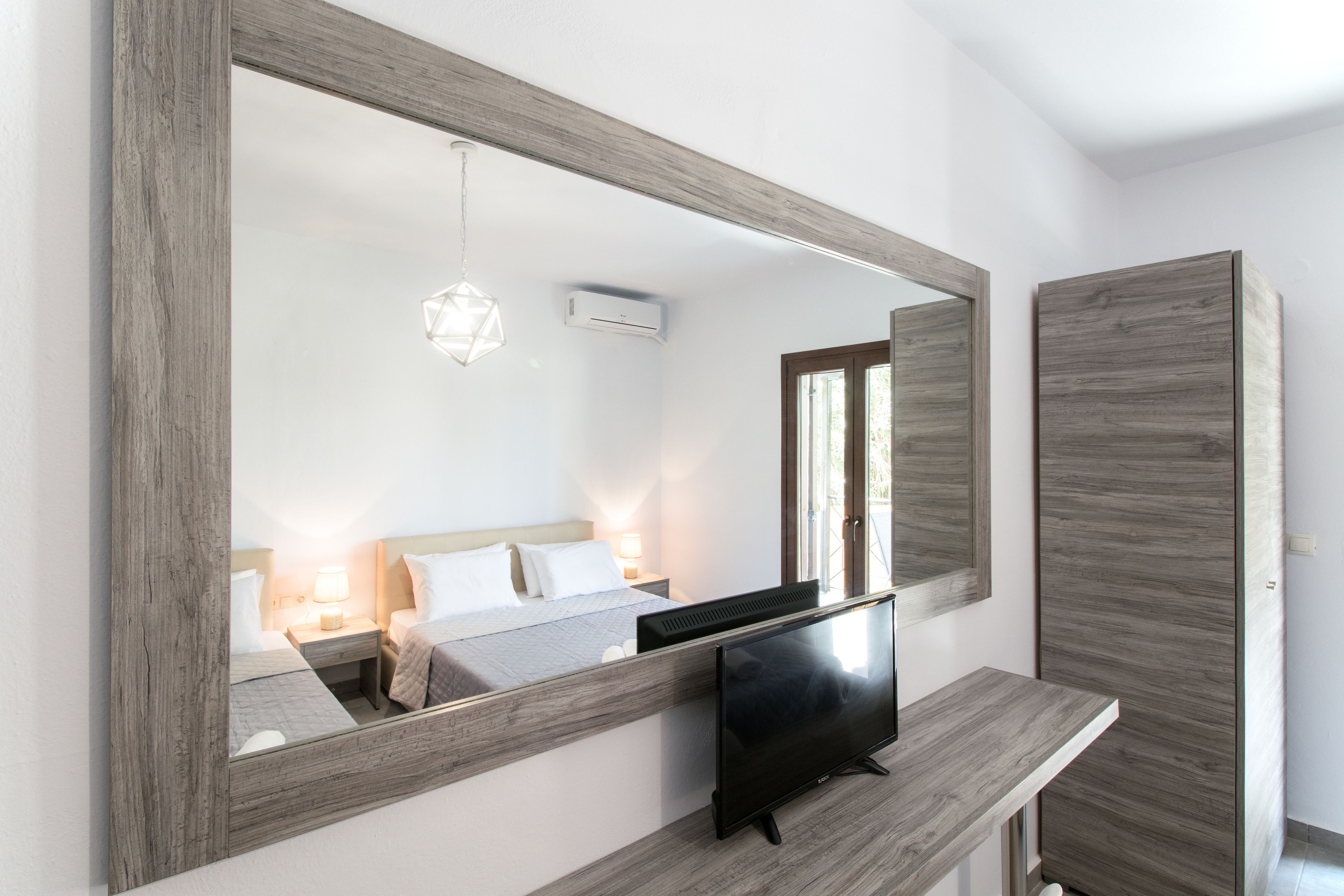 Quarto com um espelho grande, televisão, mesa de cabeceira, cama e janela.