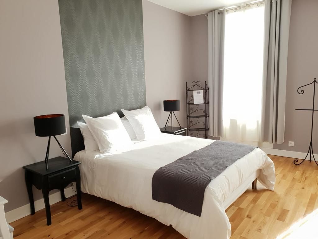 Cama bem arrumada com roupa de cama branca e manta cinza, mesa de cabeceira preta com abajur, e um suporte de metal com espelho e papéis.