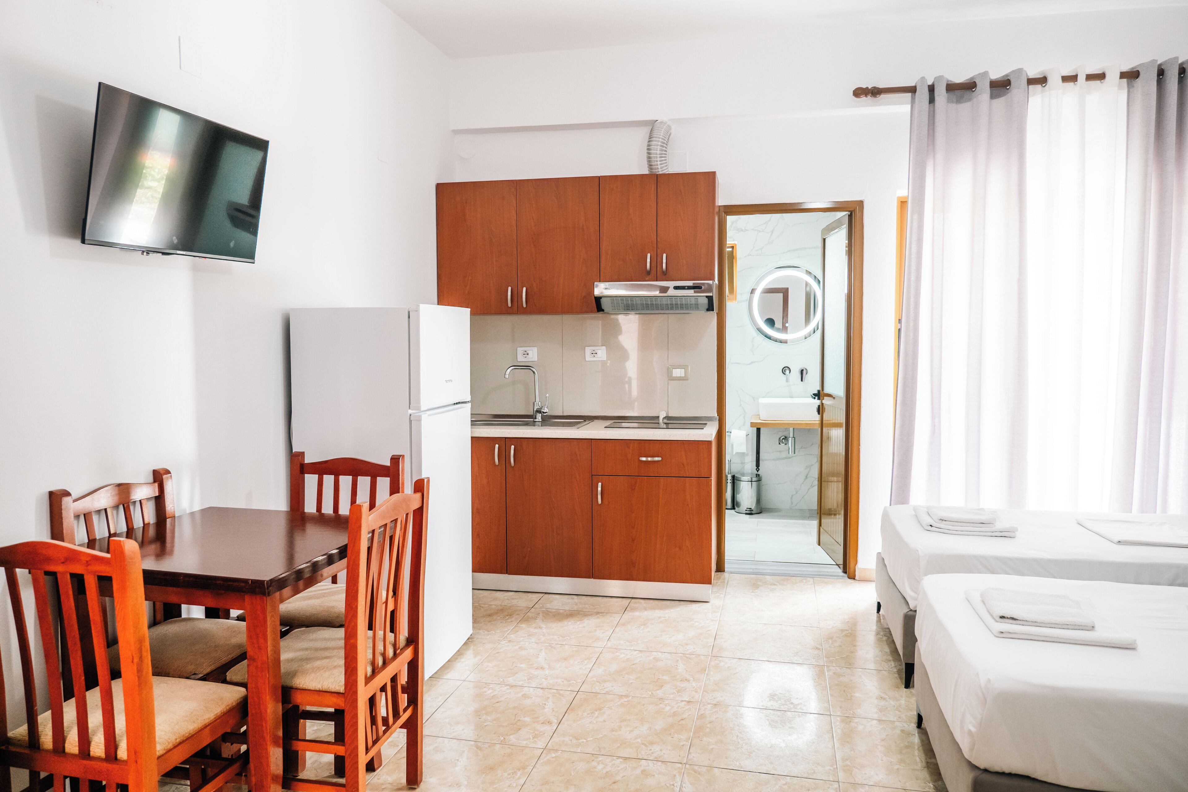 Quarto de hotel compacto com cozinha compacta, área de jantar com móveis de madeira, televisão na parede e uma cama com cortina.