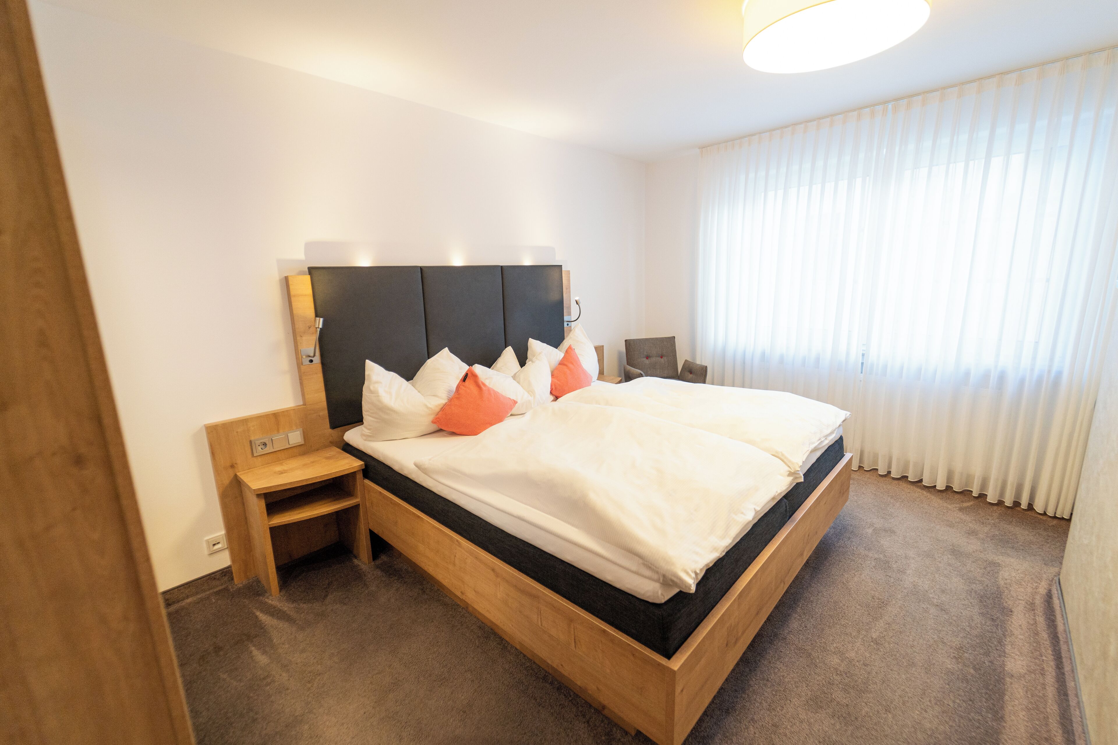 Quarto de hotel com cama de madeira, roupa de cama branca, mesa de cabeceira e uma janela grande com cortinas leves.