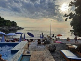 Uma área de piscina à beira-mar, com espreguiçadeiras e guarda-sóis, uma linha costeira rochosa e vista para o mar.