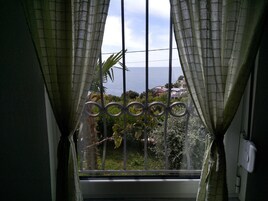 Janela com grades de metal e cortinas leves, com vista para uma paisagem costeira com vegetação e edifícios.