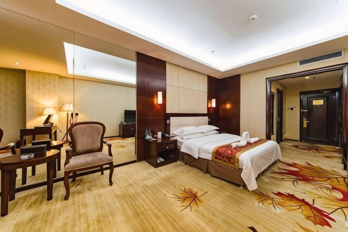 Quarto de hotel com cama, escrivaninha, cadeira, mesa de cabeceira e luminária.