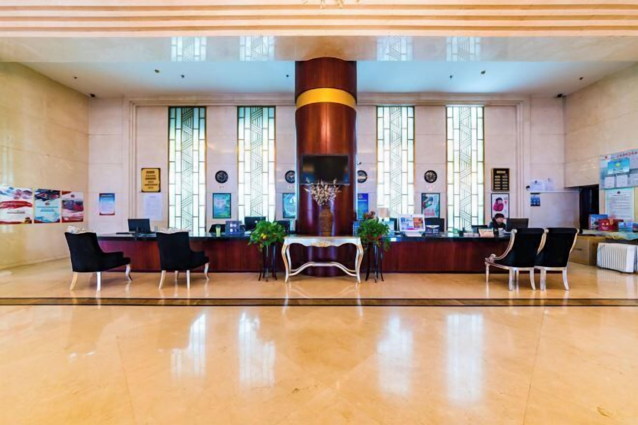 Um hall amplo com balcão de atendimento, duas cadeiras pretas, uma mesa central com plantas e um grande pilar de madeira.