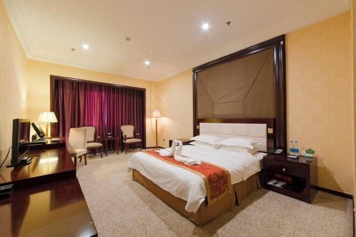Quarto de hotel com uma cama grande, uma escrivaninha com luminária, uma cadeira e uma mesa de cabeceira com telefone e garrafas.