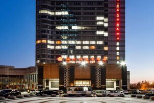 Hotel com vários andares, entrada imponente, área de estacionamento em frente e janelas iluminadas.