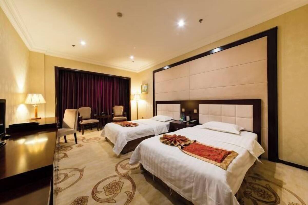 Quarto de hotel com duas camas, uma escrivaninha, uma cadeira e uma janela grande com cortinas.