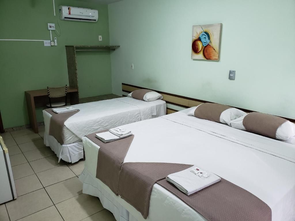 Quarto de hotel com duas camas, uma cadeira, uma mesinha e um aparelho de ar condicionado na parede.