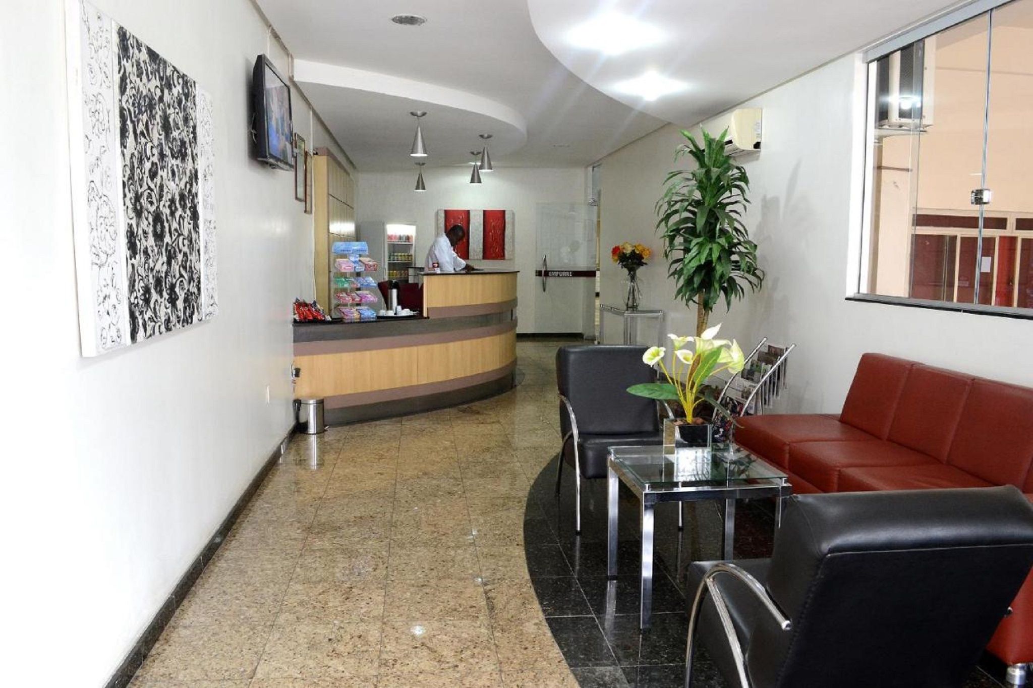 Uma área de recepção com balcão, um sofá vermelho, uma mesa de vidro com um vaso e uma planta em vaso.