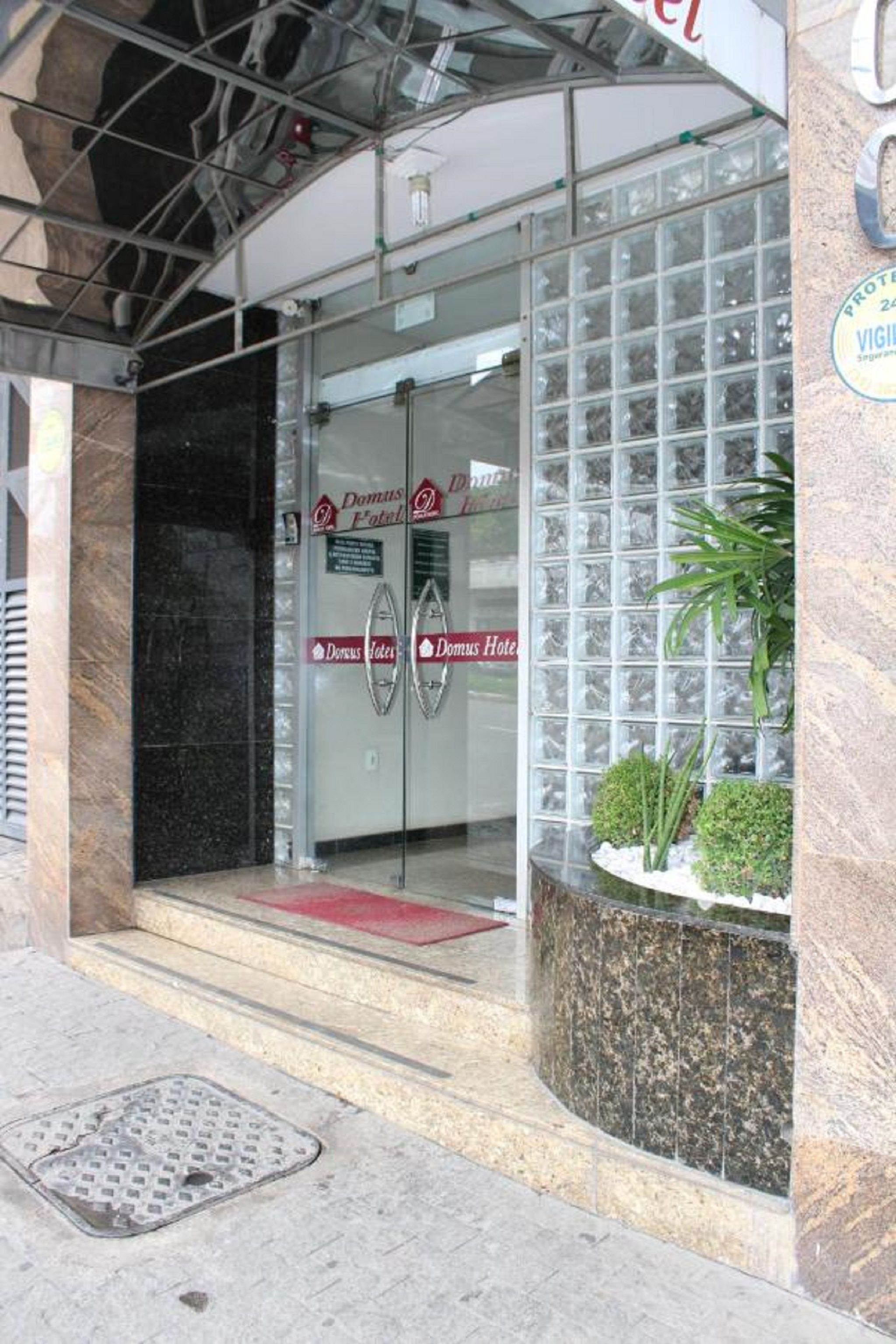 Entrada de hotel com porta de vidro, tapete vermelho e um vaso de pedra com plantas.