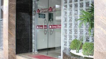 Entrada de hotel com porta de vidro, tapete vermelho e um vaso de pedra com plantas.