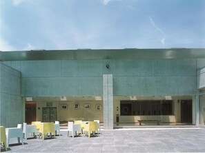 Um edifício com telhado plano, amplas janelas e área de entrada pavimentada com várias cadeiras.