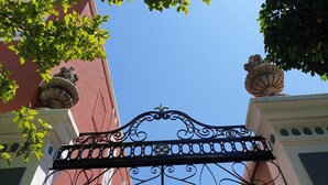 Um portão de ferro forjado com desenhos intrincados, diante de uma fachada de prédio com paredes vermelhas e brancas, sob um céu azul claro.