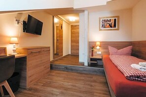 Quarto de hotel com cama, escrivaninha, cadeira, TV fixada na parede e porta para outro cômodo.