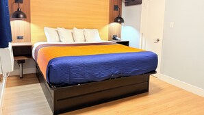 Quarto de hotel moderno com cabeceira de madeira, cama com colcha azul e laranja, duas mesas de cabeceira com abajures e uma porta.