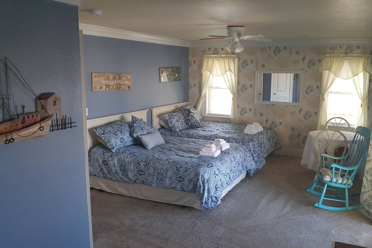 Quarto com cama, ventoinha no teto, mesinha com cadeiras e decoração náutica na parede.