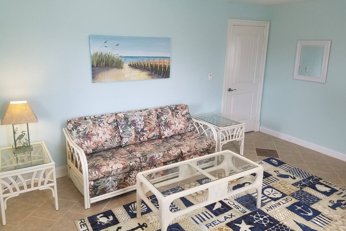 Uma sala de estar com um sofá floral, uma mesa de centro de vidro, um abajur e um quadro com uma paisagem praiana.