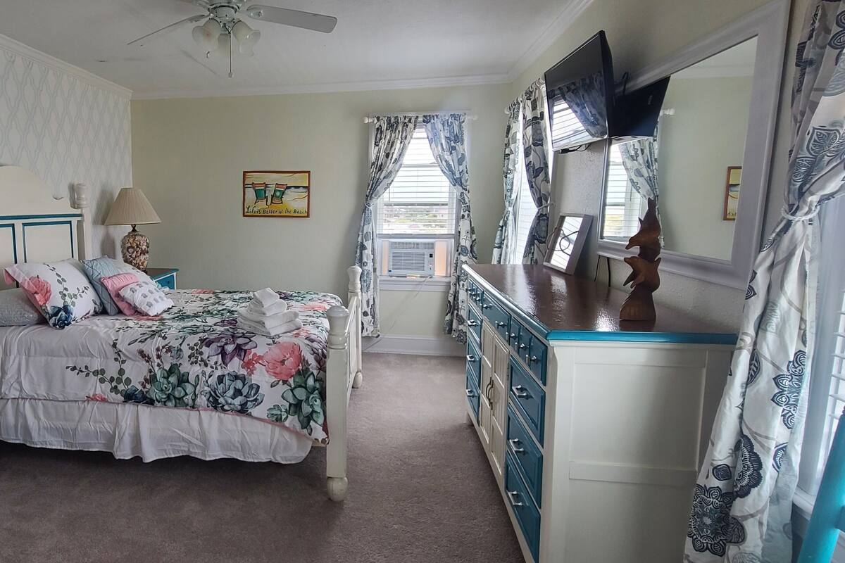 Quarto com cama, mesas de cabeceira, cômoda, ventilador de teto e janela com cortinas.