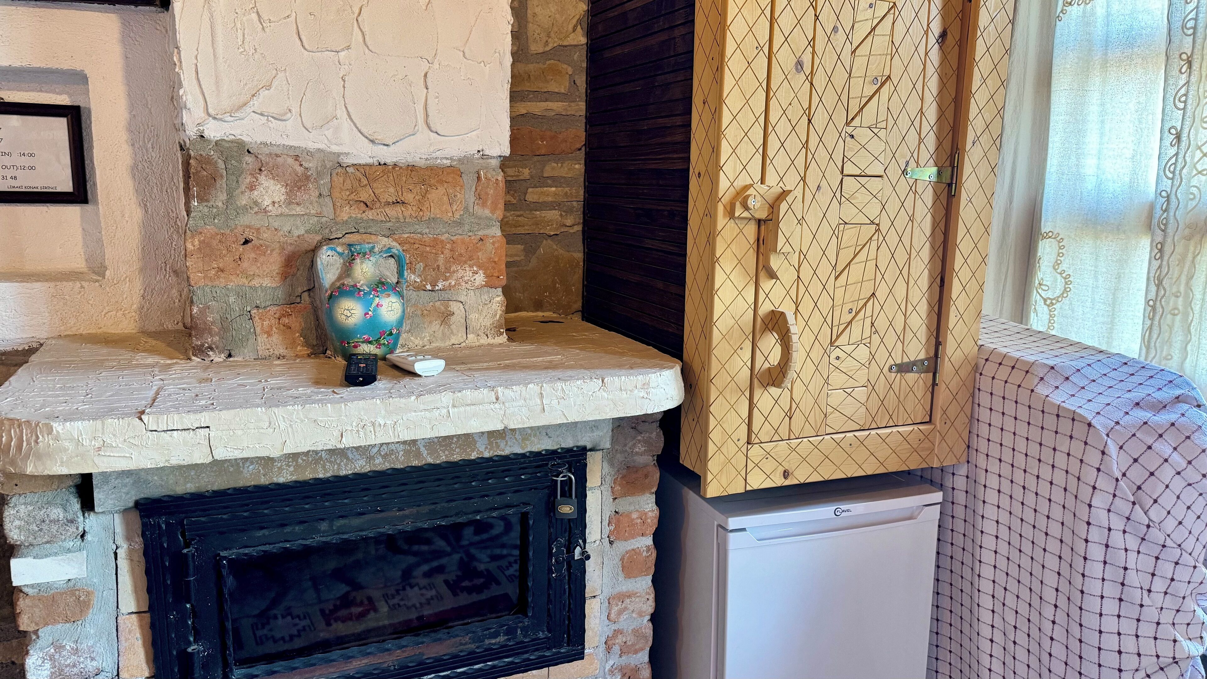 Lareira de pedra com um vaso decorativo e controle remoto, uma janela com moldura preta e um armário de madeira com porta em padrão de losangos.
