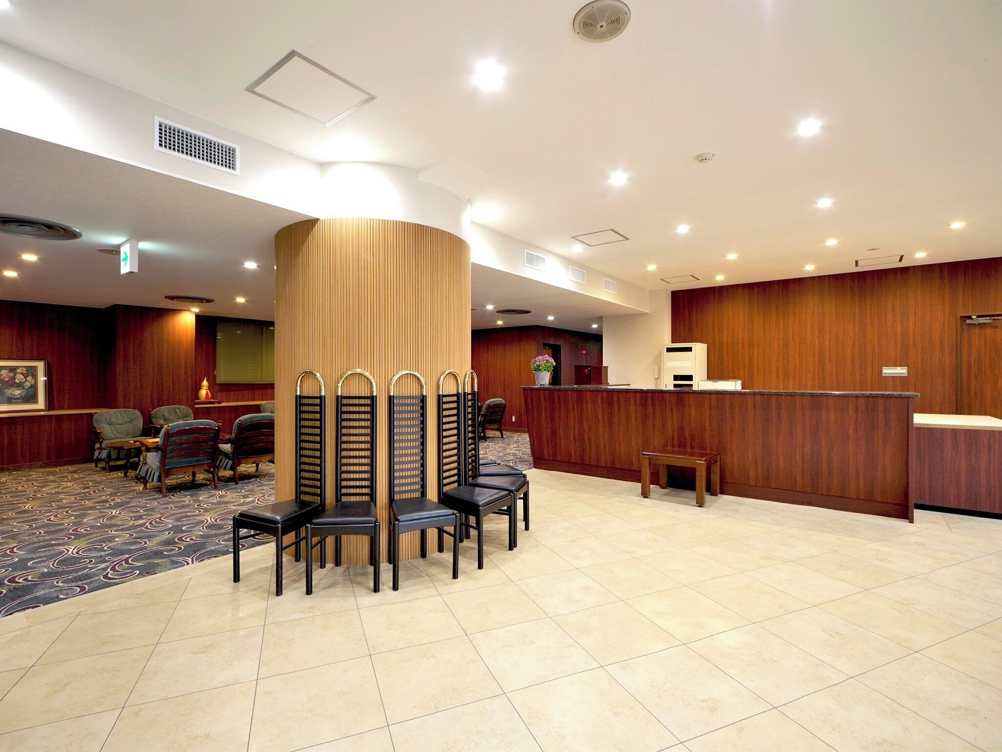 Um hall de hotel com balcão de recepção, área de estar com cadeiras, carpete estampado e um pilar grande.