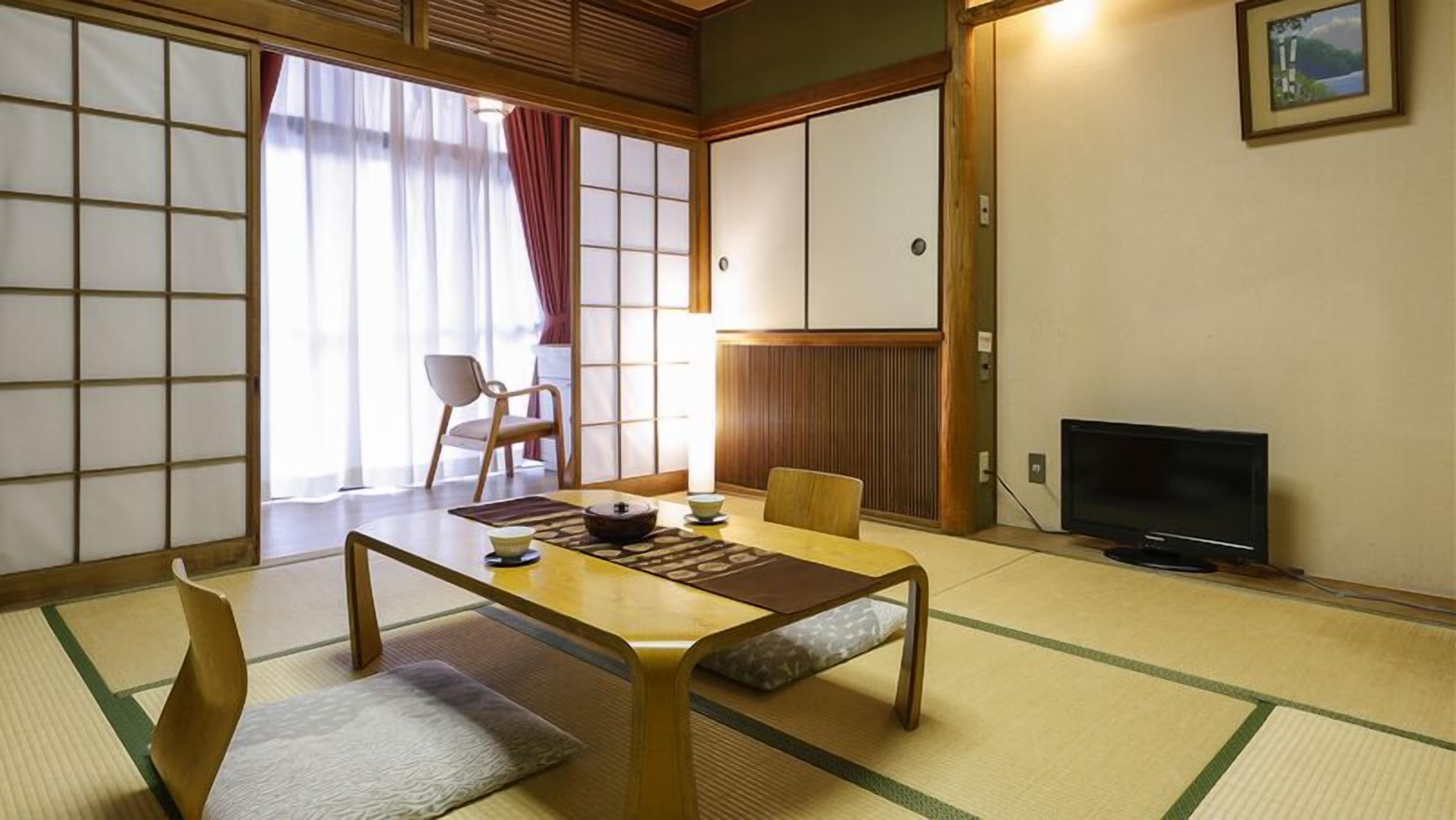 Um quarto japonês tradicional com tatame, uma mesa baixa com almofadas, uma televisão e portas deslizantes.