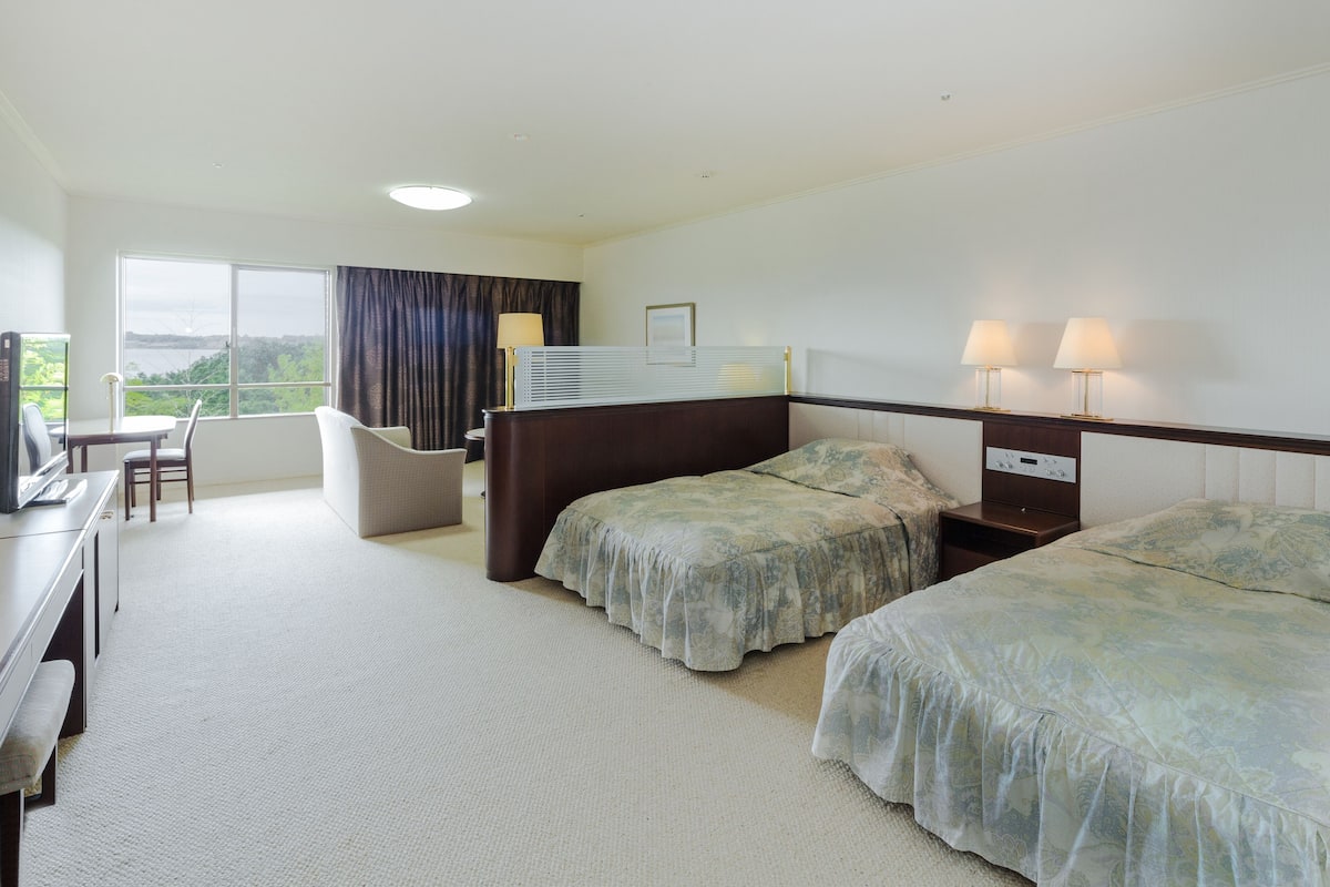 Quarto de hotel com duas camas, uma escrivaninha, uma cadeira, uma janela com vista e mesa de cabeceira com luminárias.