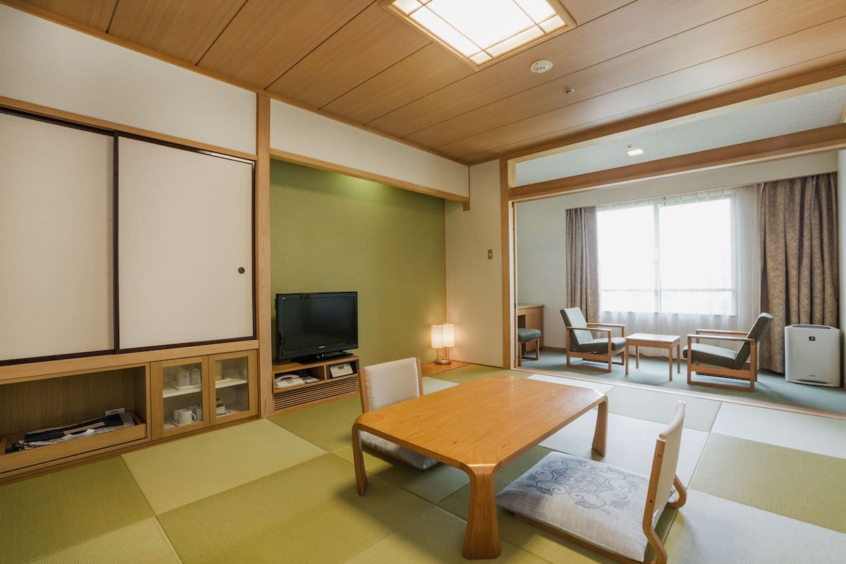Um quarto japonês tradicional com tatames, uma mesa de madeira, cadeiras, uma televisão e uma porta deslizante para outro cômodo.