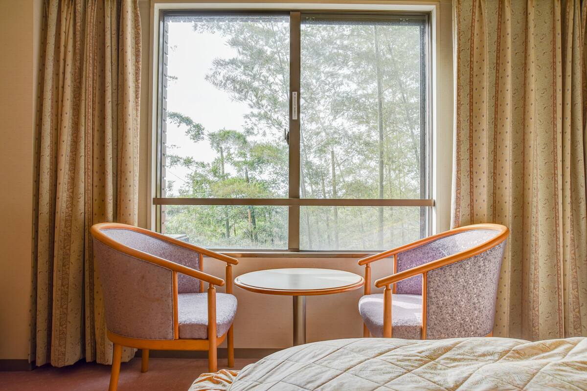 Quarto com cama, duas poltronas, uma mesa redonda pequena e uma janela grande com cortinas.