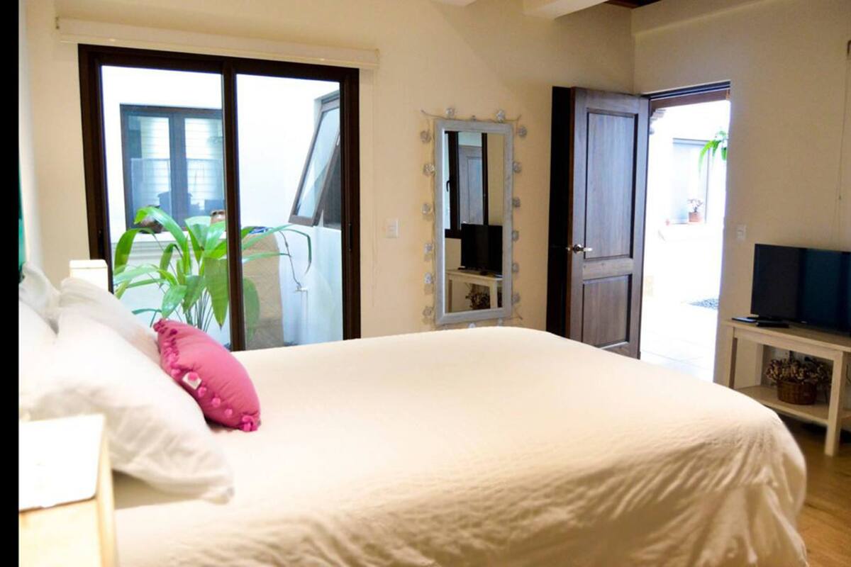 Quarto com cama grande, espelho iluminado, televisão e varanda com plantas.