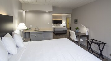 Quarto de hotel com uma cama grande, uma escrivaninha com cadeira, uma pequena cozinha e um quarto separado com uma cama.