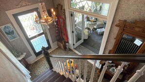 Uma escadaria com corrimão branco que leva a um quarto com lustre, papel de parede estampado e vista para área externa com mesa e cadeiras.