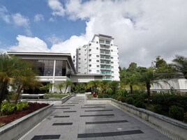 Um edifício moderno com telhado plano e grandes janelas, rodeado por área ajardinada com palmeiras e um passeio pavimentado.
