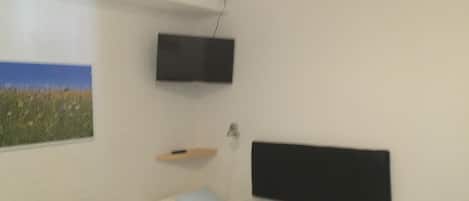 Quarto pequeno e simples com uma cama, uma TV fixada na parede e uma janela com vista para um campo.
