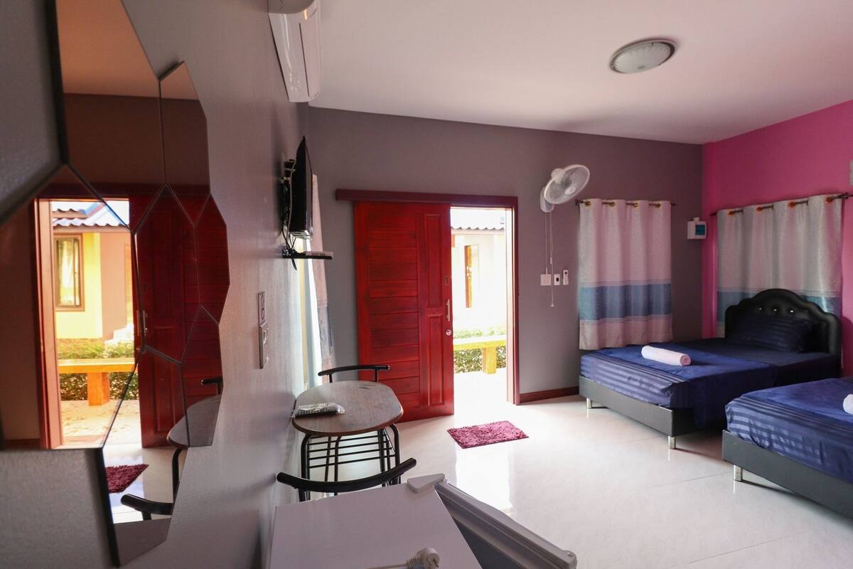 Quarto com duas camas, uma mesa, uma cadeira, uma porta vermelha e vista para o exterior.