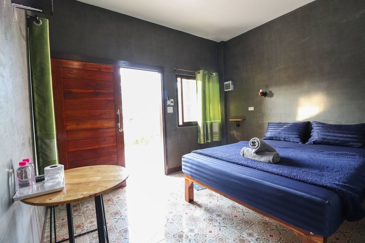 Quarto com cama, uma mesinha, porta de madeira e janela com cortinas.