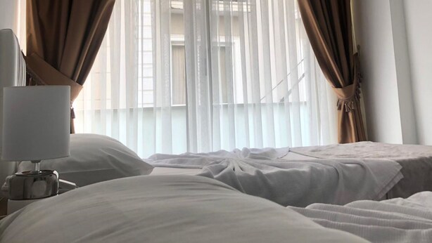 Quarto de hotel com duas camas, mesa de cabeceira com abajur e cortinas leves com cortinas marrons.