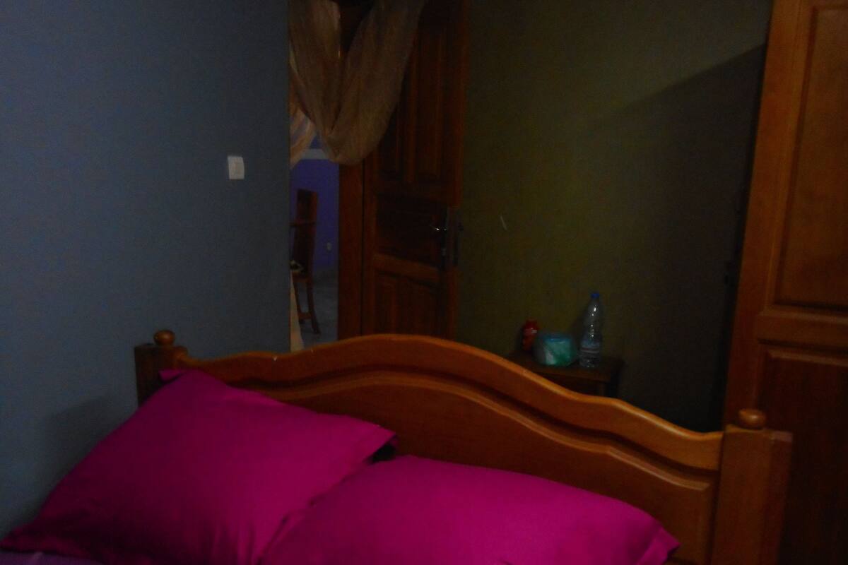 Quarto com estrutura de cama de madeira, roupa de cama roxa e uma mesa de cabeceira com uma garrafa.