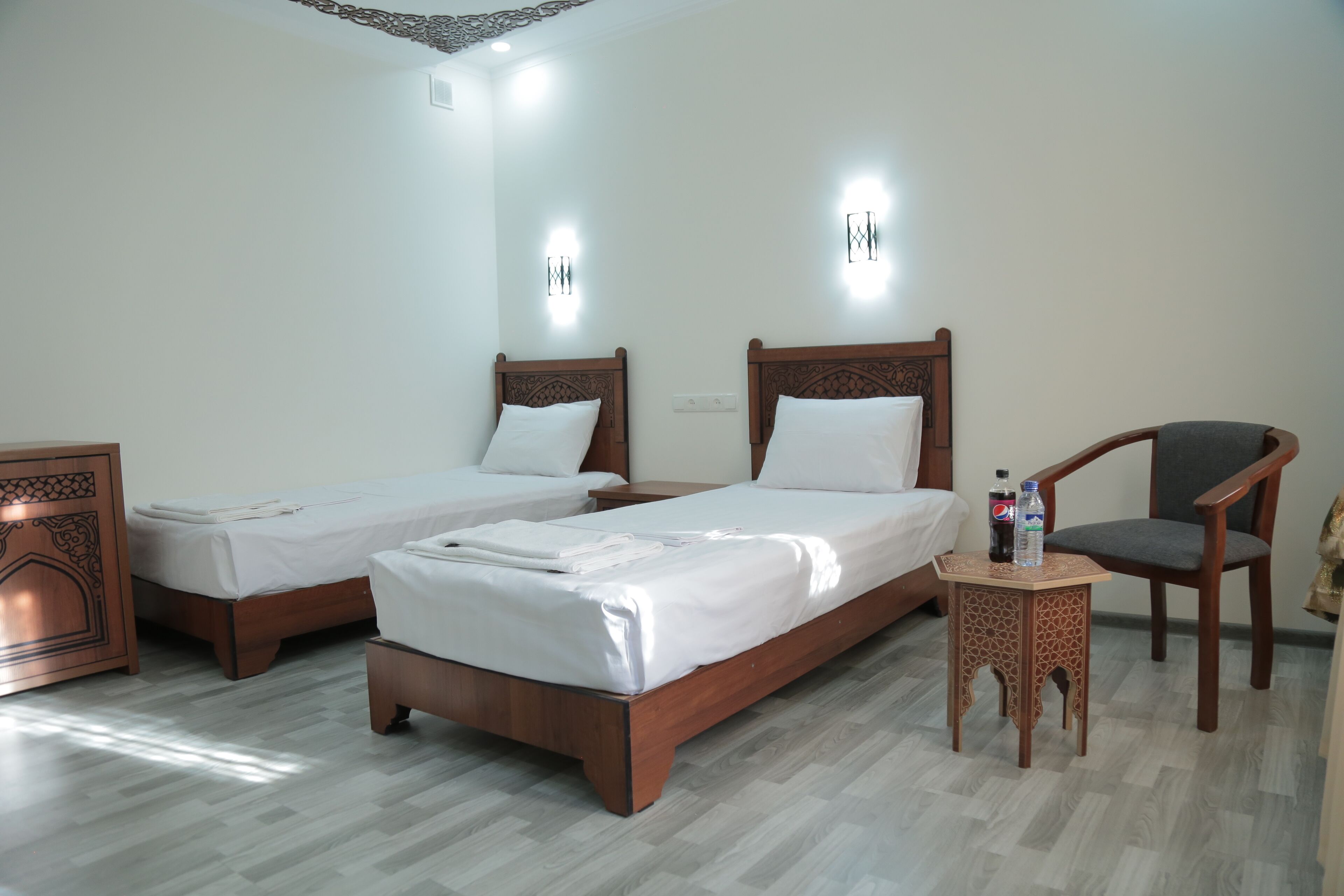 Quarto de hotel com duas camas de madeira, uma pequena mesa de madeira com uma garrafa e uma garrafa de água, e uma cadeira.
