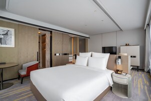 Quarto de hotel com uma cama grande, uma escrivaninha, uma cadeira, um guarda-roupa e uma mesa de cabeceira com abajur.