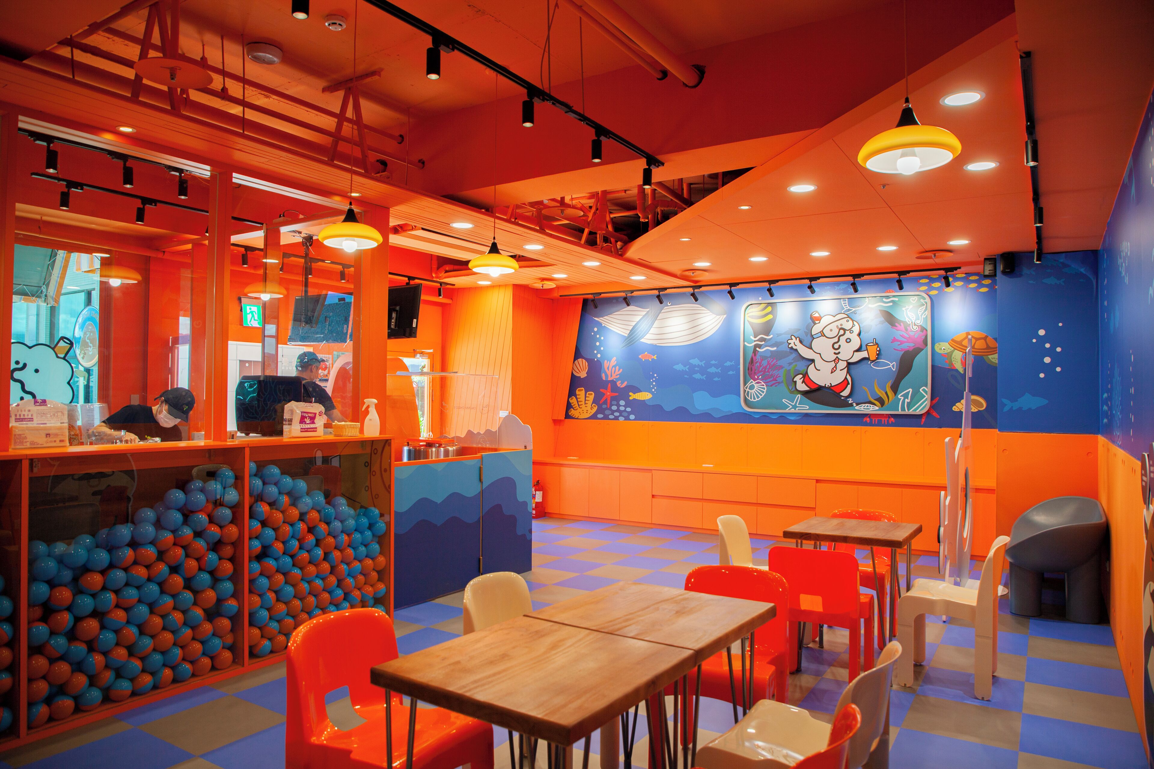 Um restaurante bem iluminado com decoração laranja e azul, com um balcão exibindo bolas azuis e laranjas, e um mural de um personagem de desenho animado.