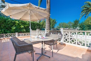 Um pátio externo com mesa redonda e duas cadeiras de vime, um guarda-sol bege e um cercado decorativo.