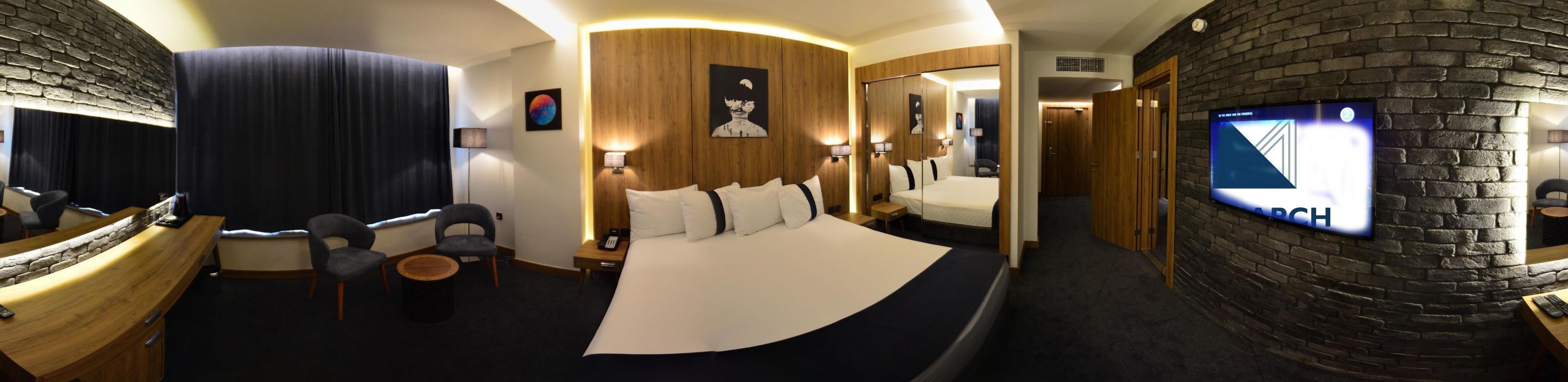 Quarto de hotel com cama curva, uma escrivaninha com cadeira, televisão e parede de pedra.