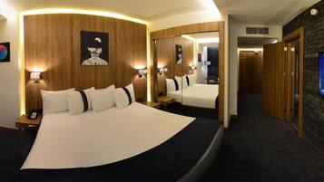 Quarto de hotel com cama curva, uma escrivaninha com cadeira, televisão e parede de pedra.