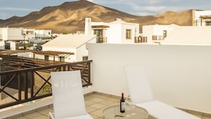 Terraço com duas poltronas brancas e uma mesa pequena, com vista para uma paisagem montanhosa e edifícios brancos.
