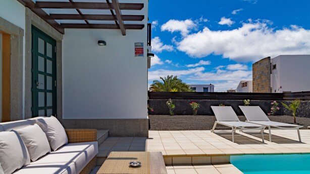 Uma área à beira da piscina com um sofá branco, duas espreguiçadeiras e uma mesa de vime.