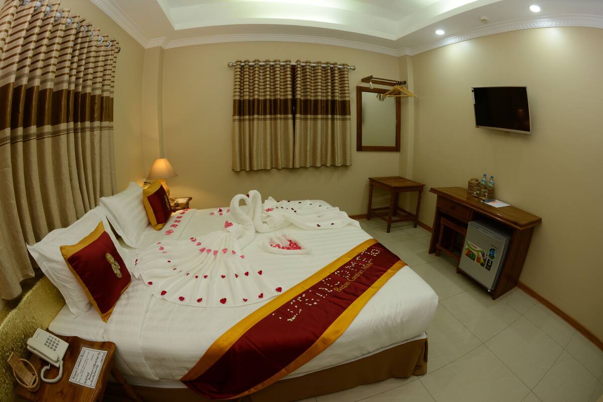 Quarto de hotel com uma cama bem arrumada, mesa de cabeceira com abajur, pequena escrivaninha com espelho, televisão e um frigorífico.