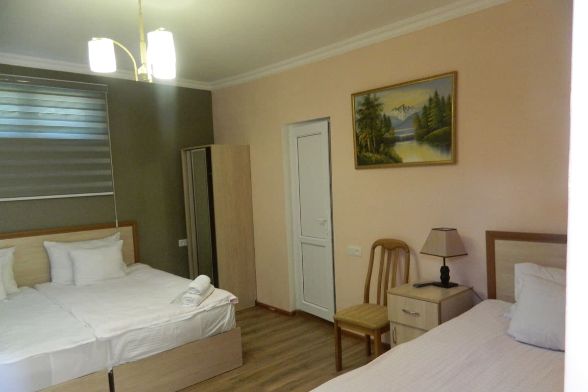 Quarto de hotel com duas camas, uma cadeira, uma mesa de cabeceira com abajur, guarda-roupa, uma porta e um quadro na parede.