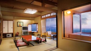 Um quarto japonês tradicional com tatames, uma mesa baixa com almofadas, uma televisão e uma grande janela com vista para o mar.