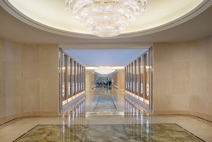 Um corredor luxuoso com piso de mármore refletivo, um lustre imponente e paredes espelhadas.